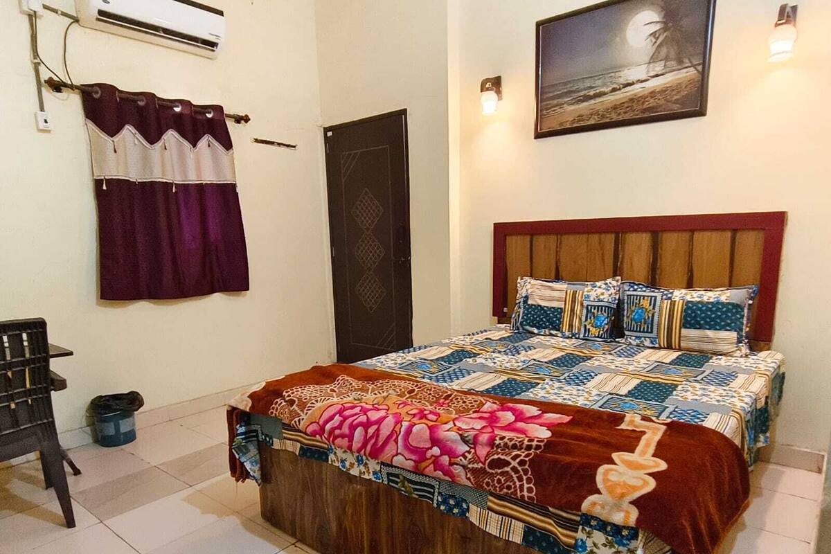 Quarto com cama, uma cadeira, ar condicionado, porta, quadro na parede e tapete vermelho.