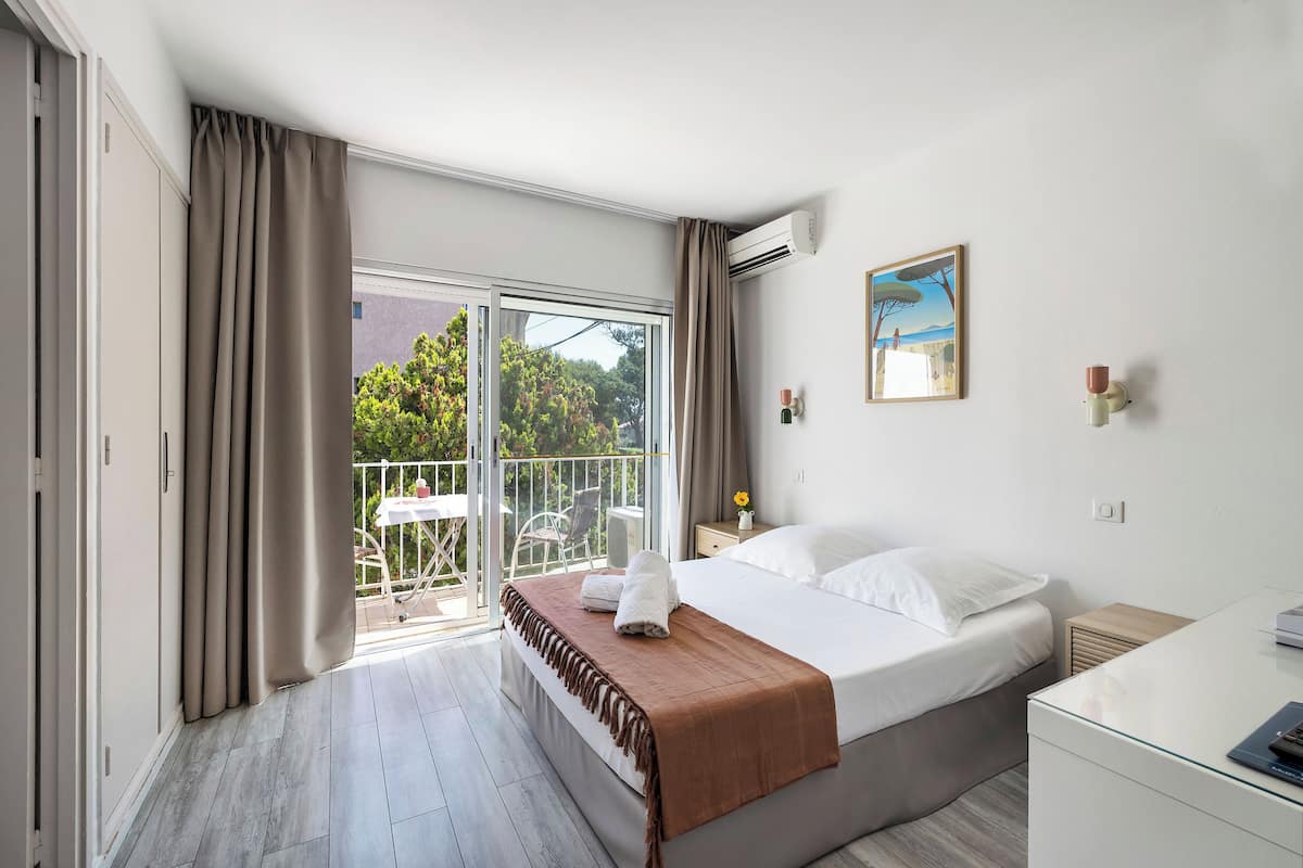 Quarto de hotel com cama, escrivaninha, cadeira, varanda com mesa e cadeiras, e um quadro na parede.