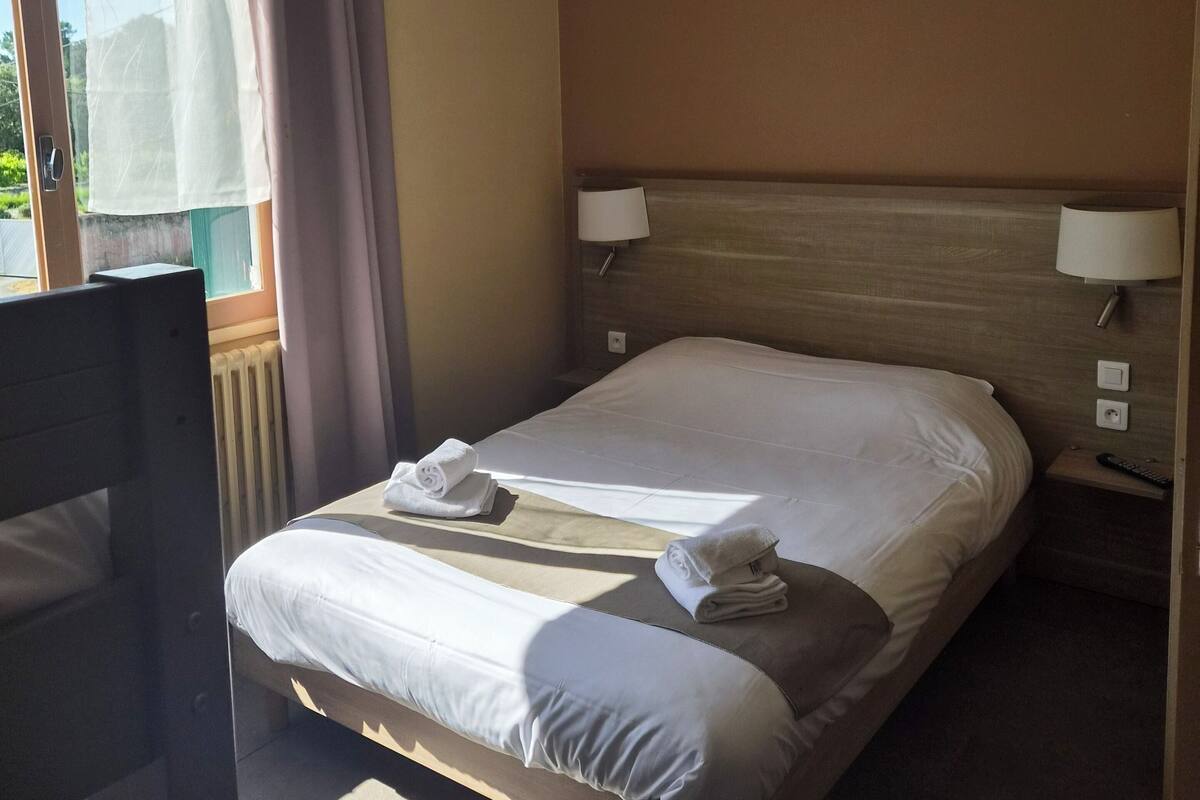 Quarto de hotel com cama, duas luminárias de cabeceira, uma cadeira e uma janela com cortinas.