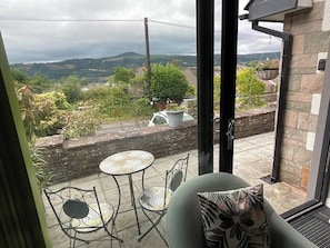 Varanda com vista para uma paisagem montanhosa, um muro de pedra e uma mesa de metal com cadeiras.