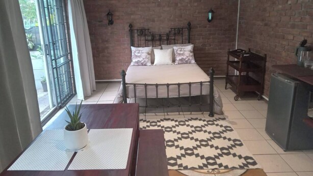 Quarto com cama, escrivaninha, cadeira, planta em vaso, tapete estampado e parede de tijolos.