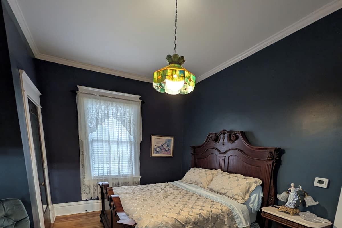 Quarto com cama, cadeira, mesa de cabeceira, luminária e janela com cortinas.