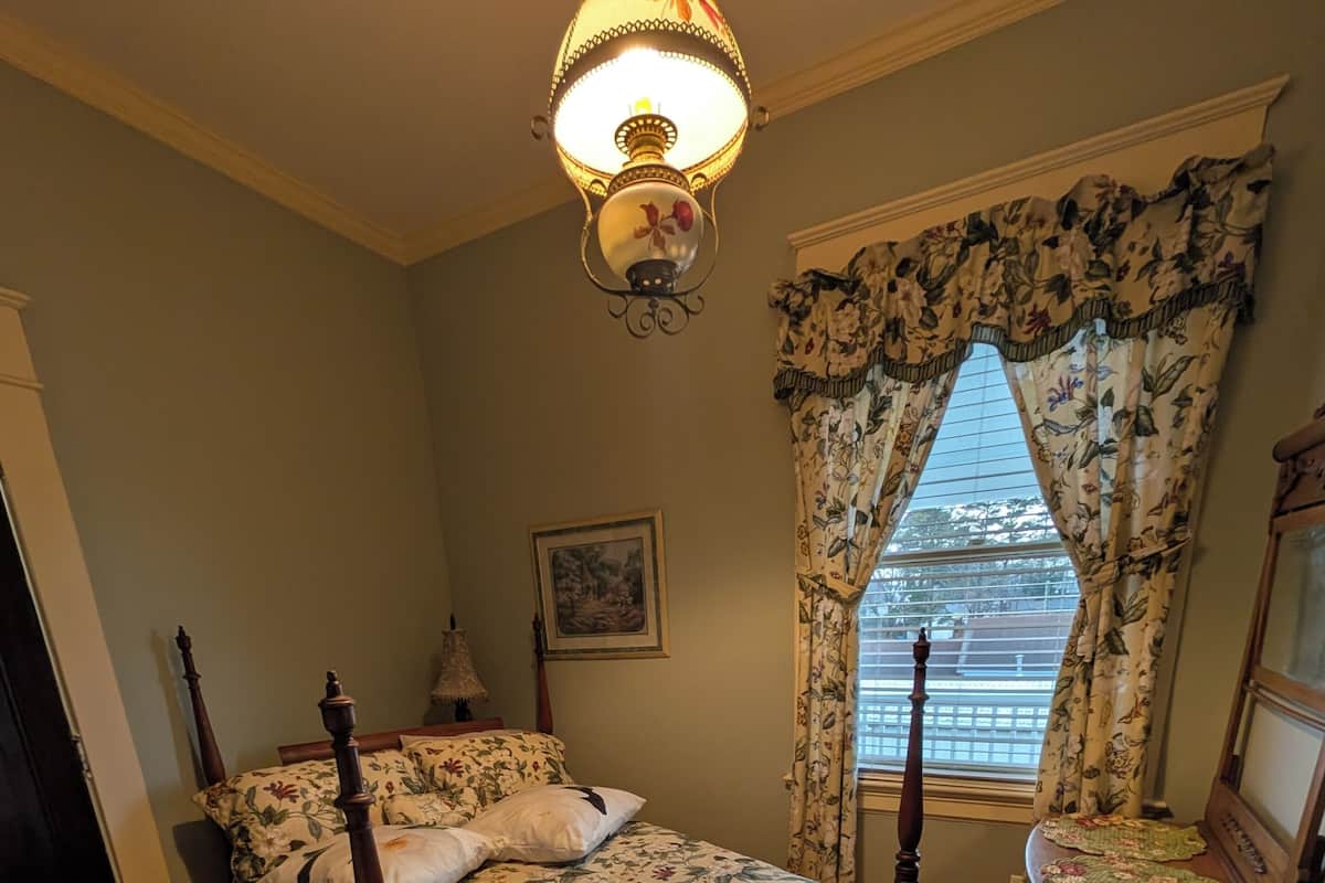 Quarto com cama de dossel, mesa de cabeceira, cômoda, janela com cortinas florais e luminária pendente.