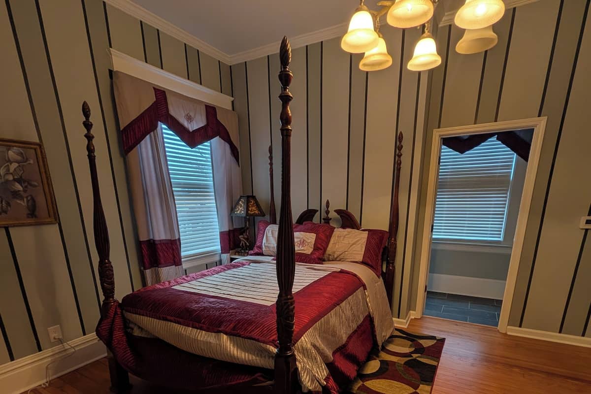 Uma cama com dossel e cortina, um candelabro, piso de madeira e um tapete estampado.