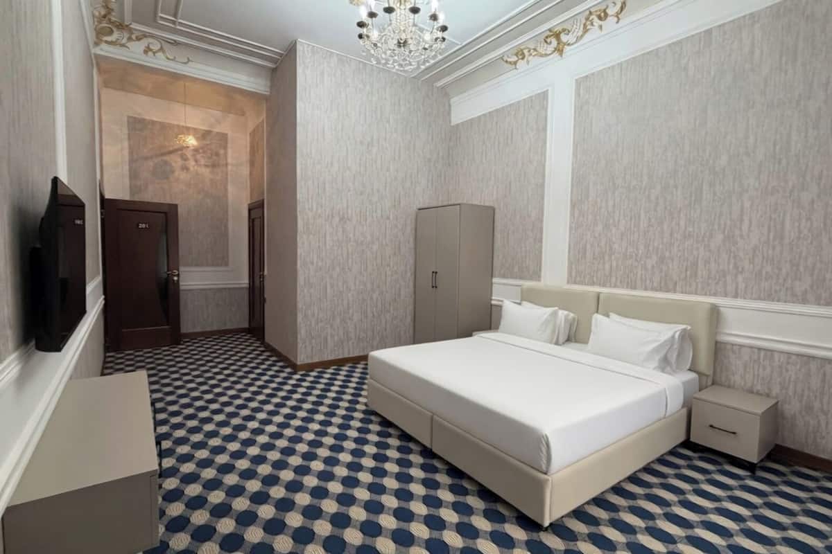 Quarto de hotel com piso xadrez, cama com lençóis brancos, guarda-roupa, mesa de cabeceira e televisão.