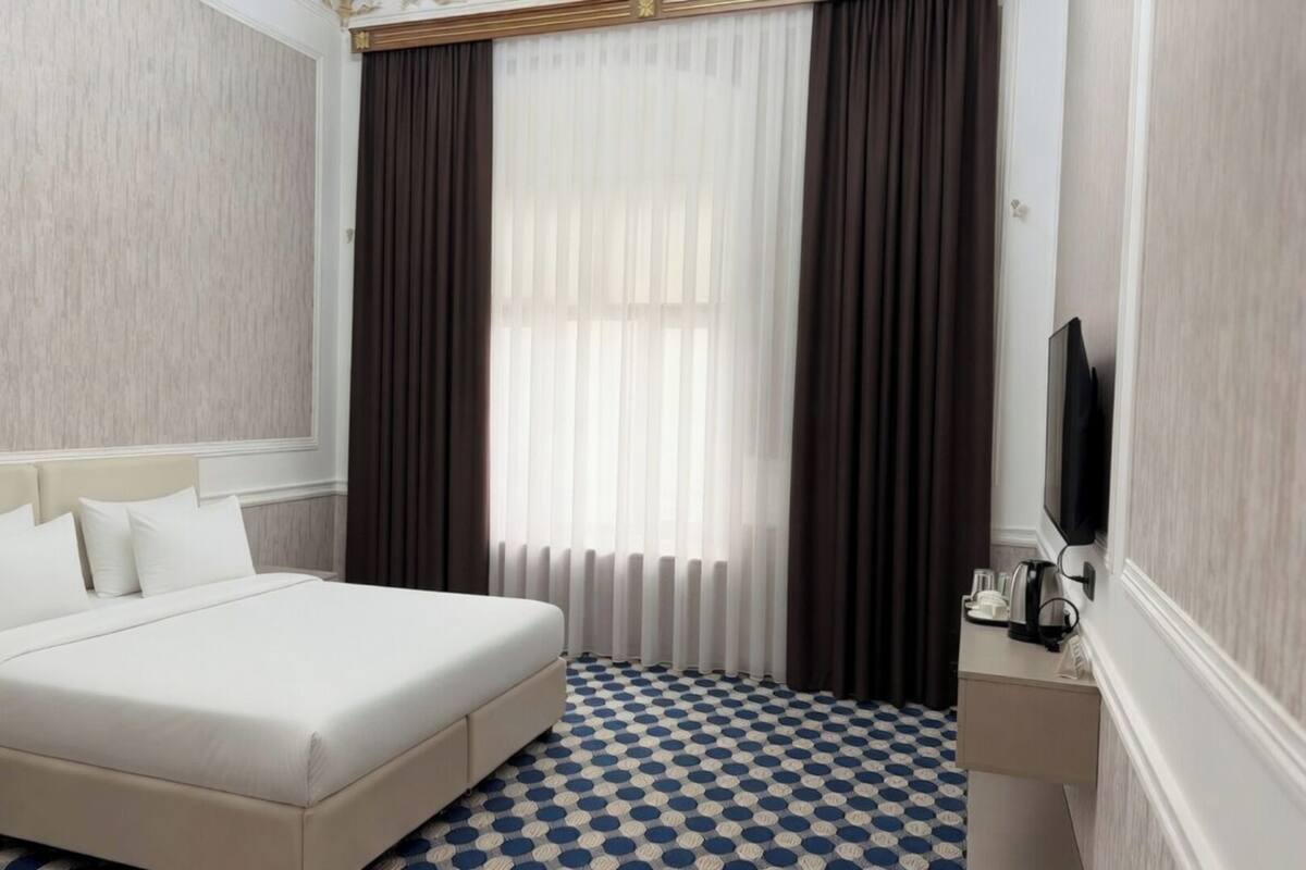 Quarto de hotel com cama, mesa de cabeceira, televisão e janela com cortinas.