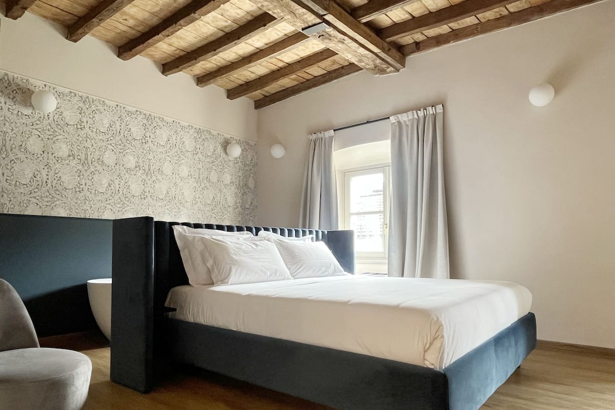 Quarto com cama, uma cadeira, janela com cortinas e teto de madeira.