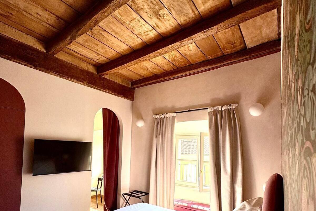 Quarto com teto de madeira, cama com lençóis brancos, televisão na parede e janela com cortinas.