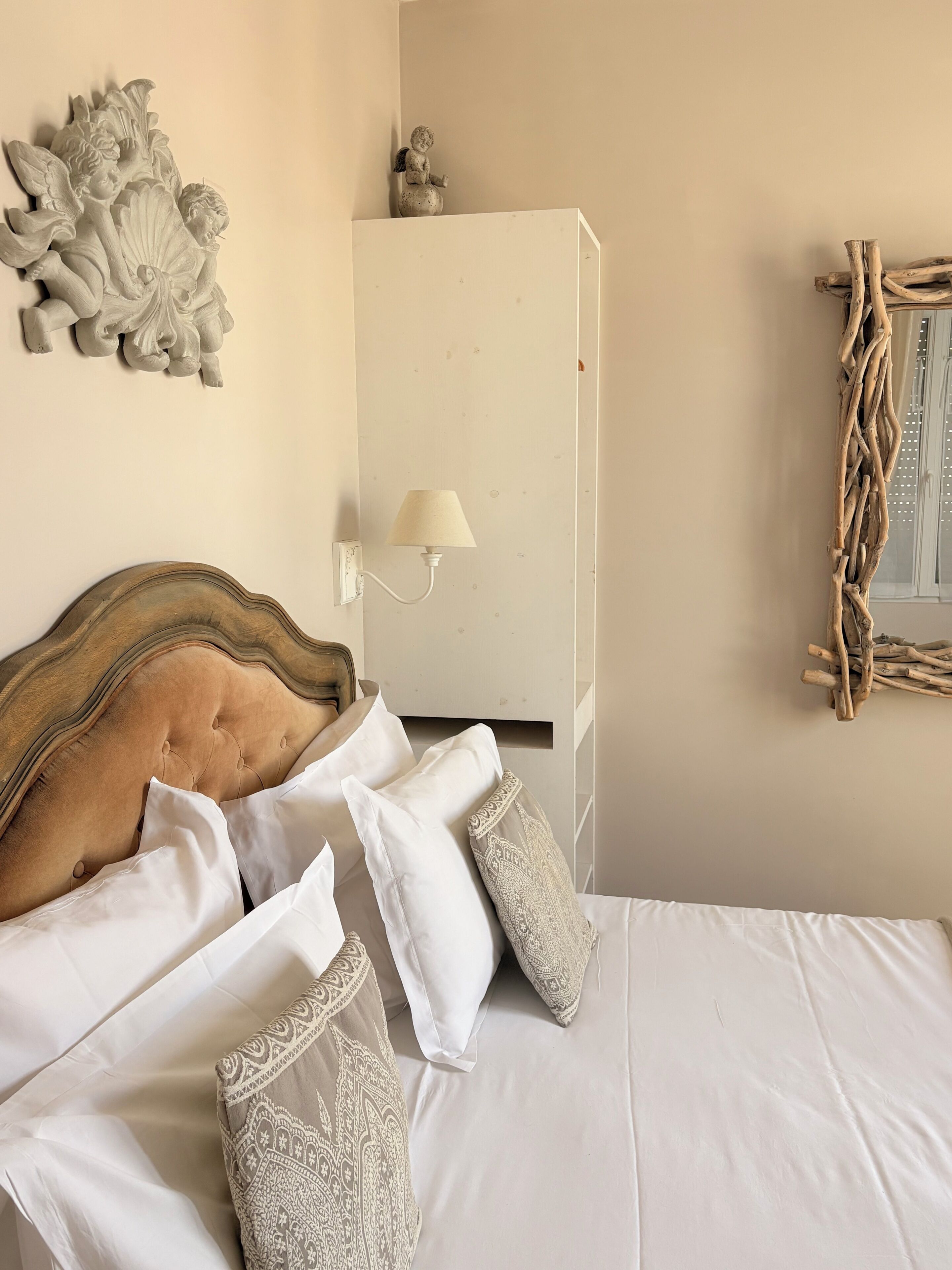 Cama bem arrumada com lençóis brancos e almofadas estampadas, luminária fixada na parede e uma escultura decorativa de anjo.