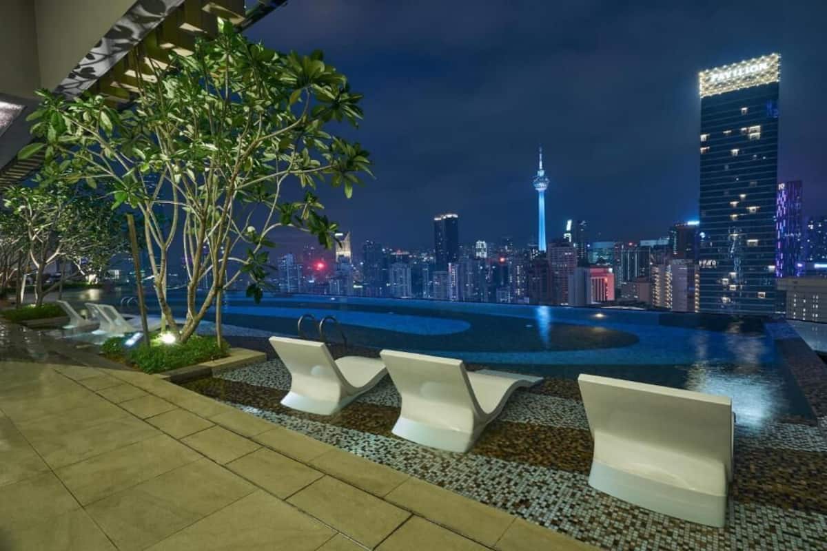 Área de piscina externa com espreguiçadeiras, um horizonte urbano ao fundo e um edifício moderno com sinalização iluminada.