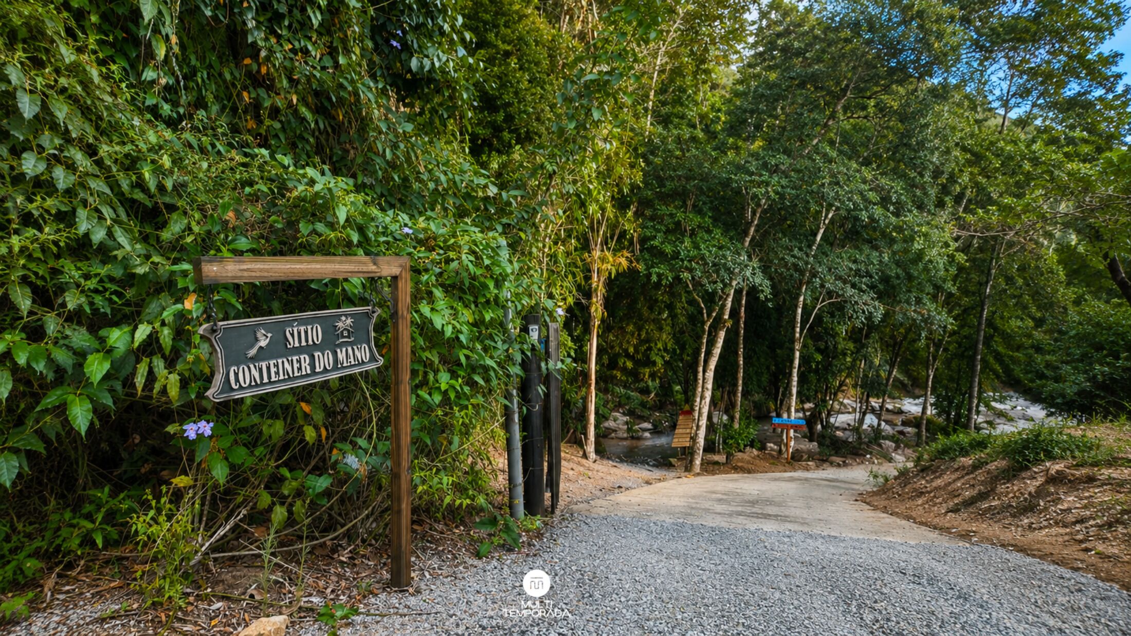 Uma trilha de cascalho serpenteia por uma área arborizada, com uma placa que diz 'SITIO CONTEINER DO MANO'.