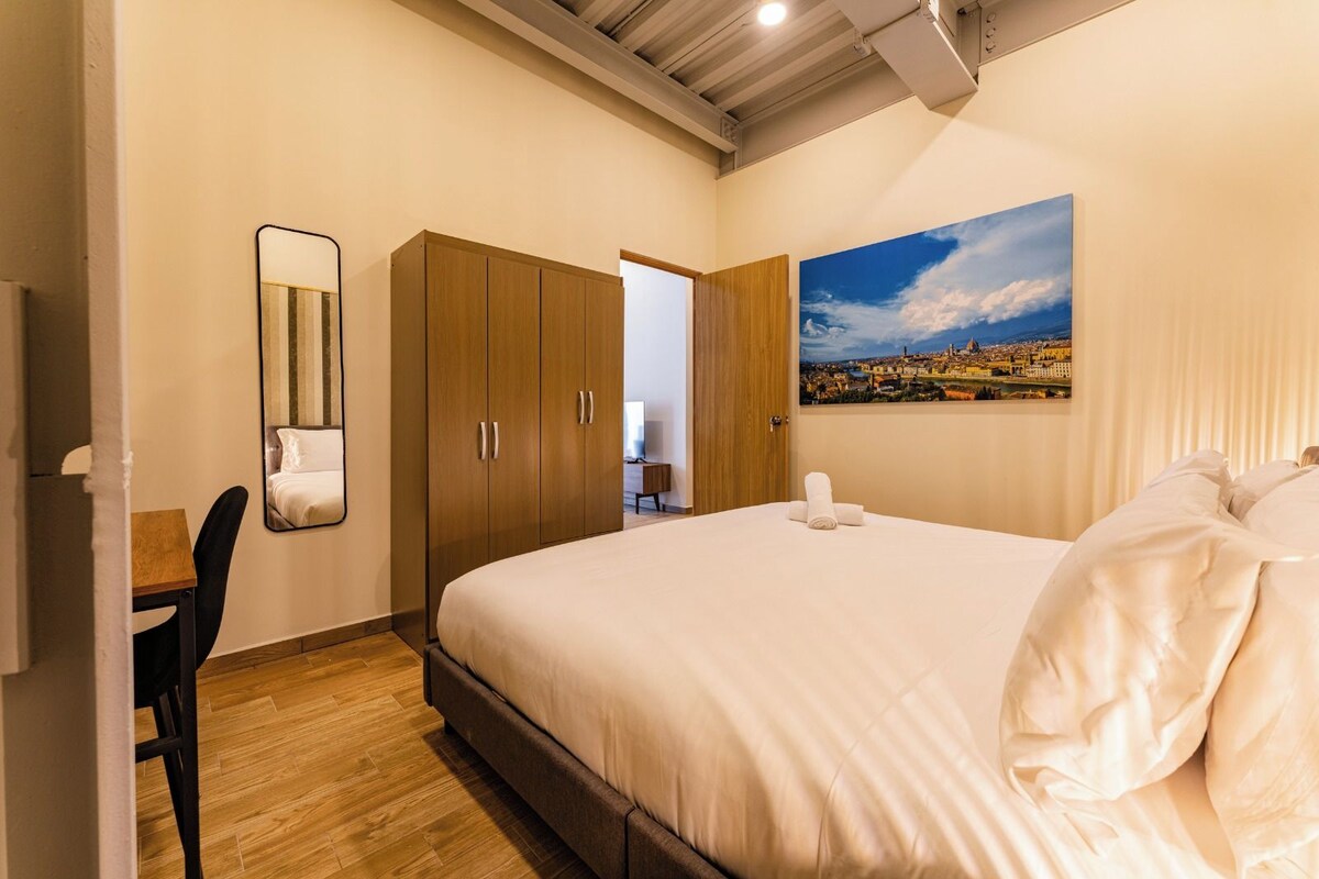 Um quarto com uma cama, uma escrivaninha, um espelho e uma porta que dá para outro cômodo.