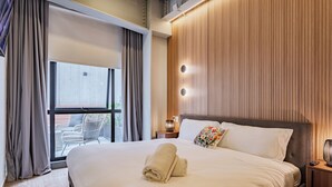 Quarto de hotel com uma cama grande, janela com cortinas, painel de parede de madeira e uma pequena mesa com abajur.