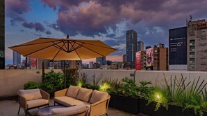 Terraço no telhado com móveis de vime, um guarda-sol e plantas em vasos, com a silhueta de uma cidade ao fundo.