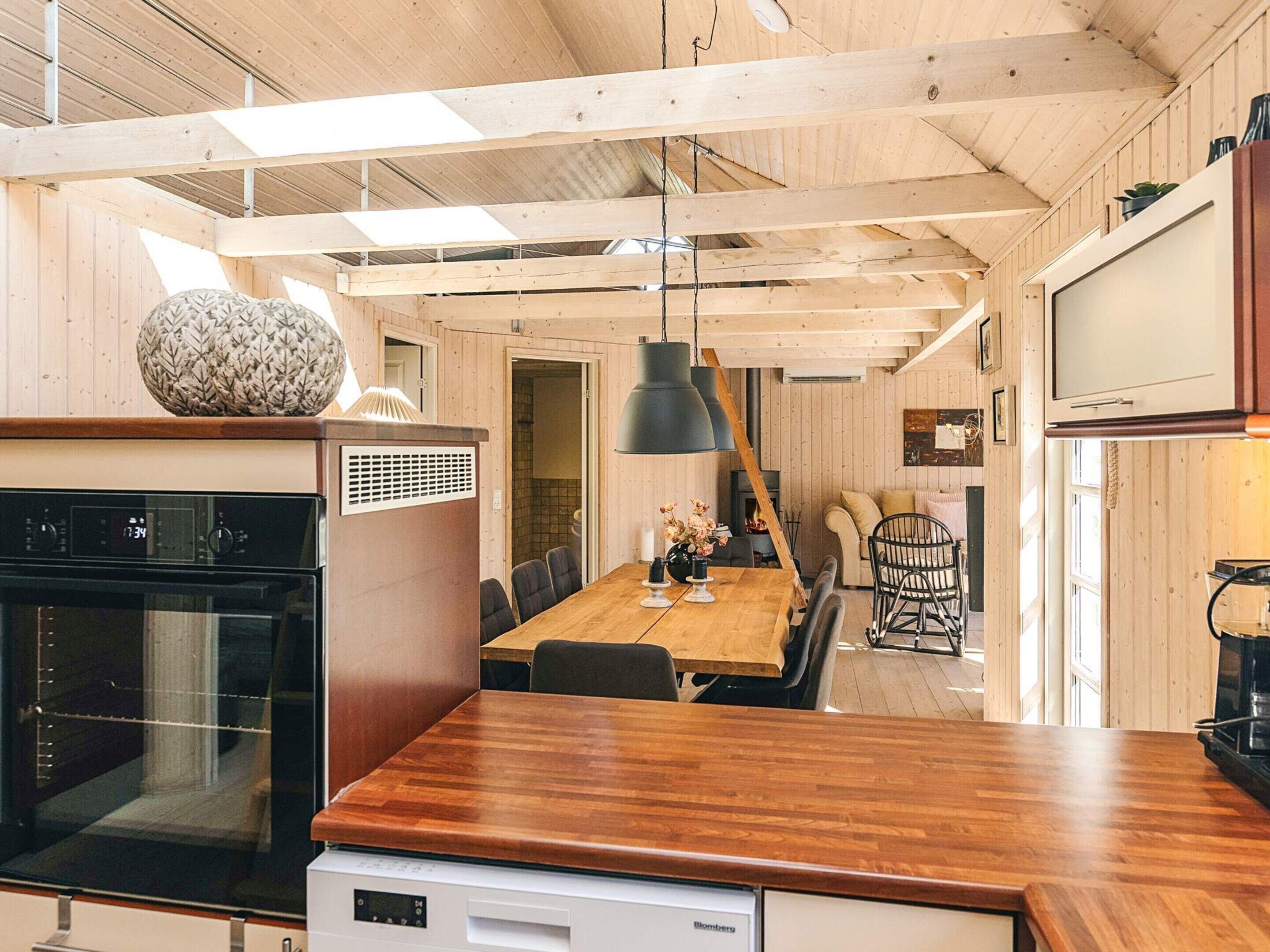Uma cozinha moderna com bancada de madeira, forno embutido e máquina de lavar louça. Há um espaço de refeições com mesa e cadeiras de madeira, e um lustre.