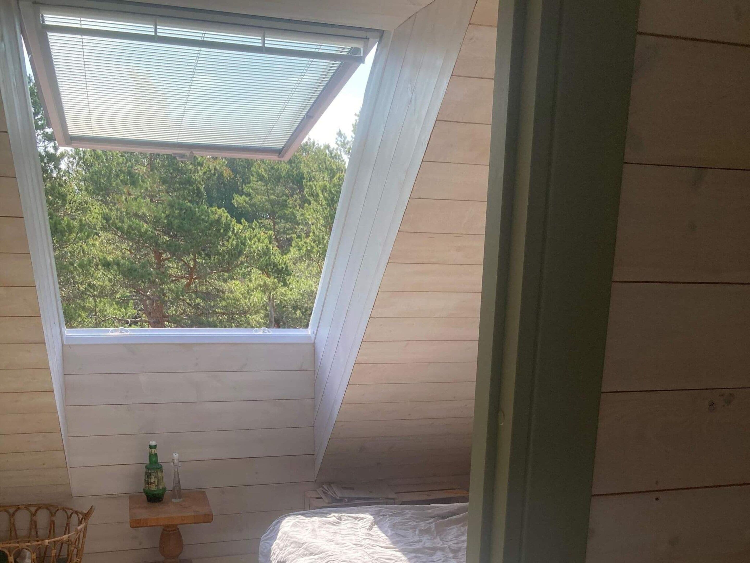 Quarto com teto inclinado, janela com vista para árvores, uma cama, uma mesinha com uma garrafa e uma cadeira.