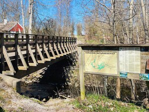 Uma ponte de madeira sobre um riacho, com uma placa de sinalização exibindo informações sobre a Reserva Natural de Verkeans.