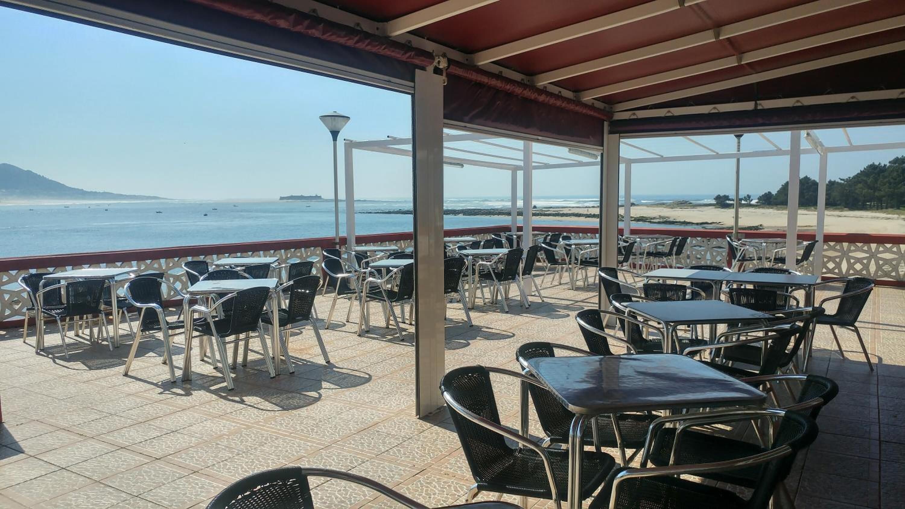 Pátio externo com mesas e cadeiras, com vista para uma praia e oceano.