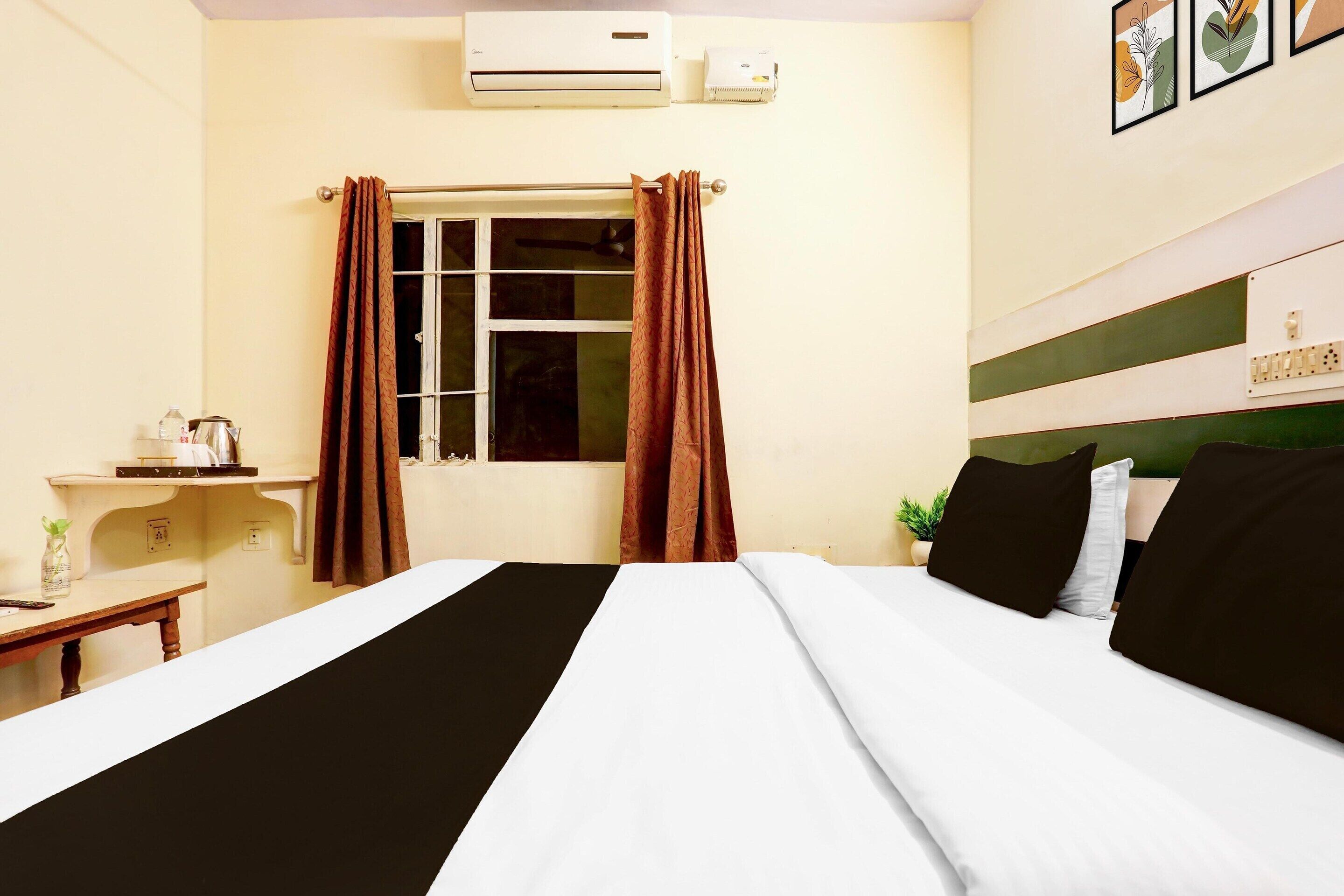 Quarto de hotel com cama, janela com cortinas, ar condicionado, mesa pequena com chaleira e prateleira fixada na parede.