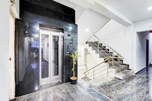 Um lobby moderno com piso de mármore, um vaso de plantas, um elevador de vidro e uma escada com corrimãos de metal.