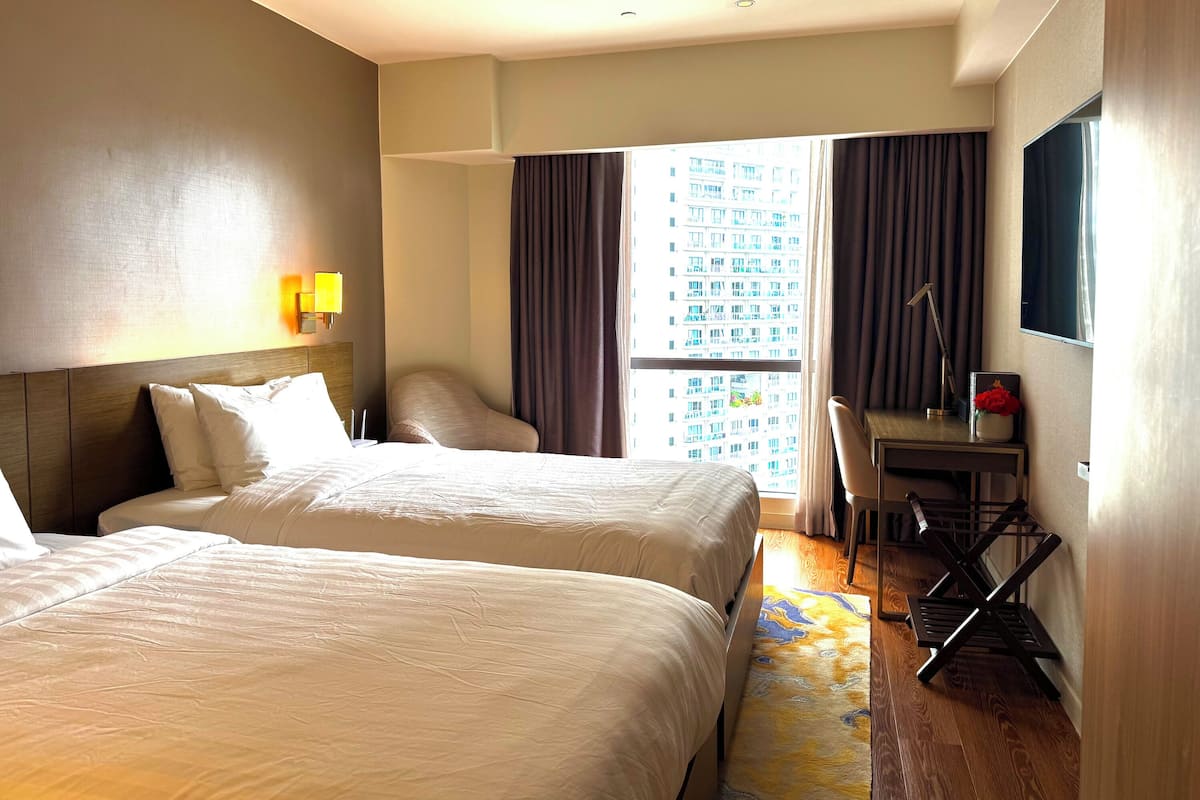 Quarto de hotel com duas camas, uma escrivaninha, uma cadeira, uma televisão e uma janela com cortinas.