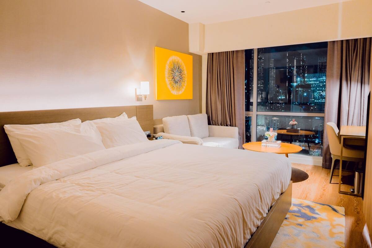 Quarto de hotel com uma cama grande, um sofá, uma mesinha e vista para a cidade à noite.