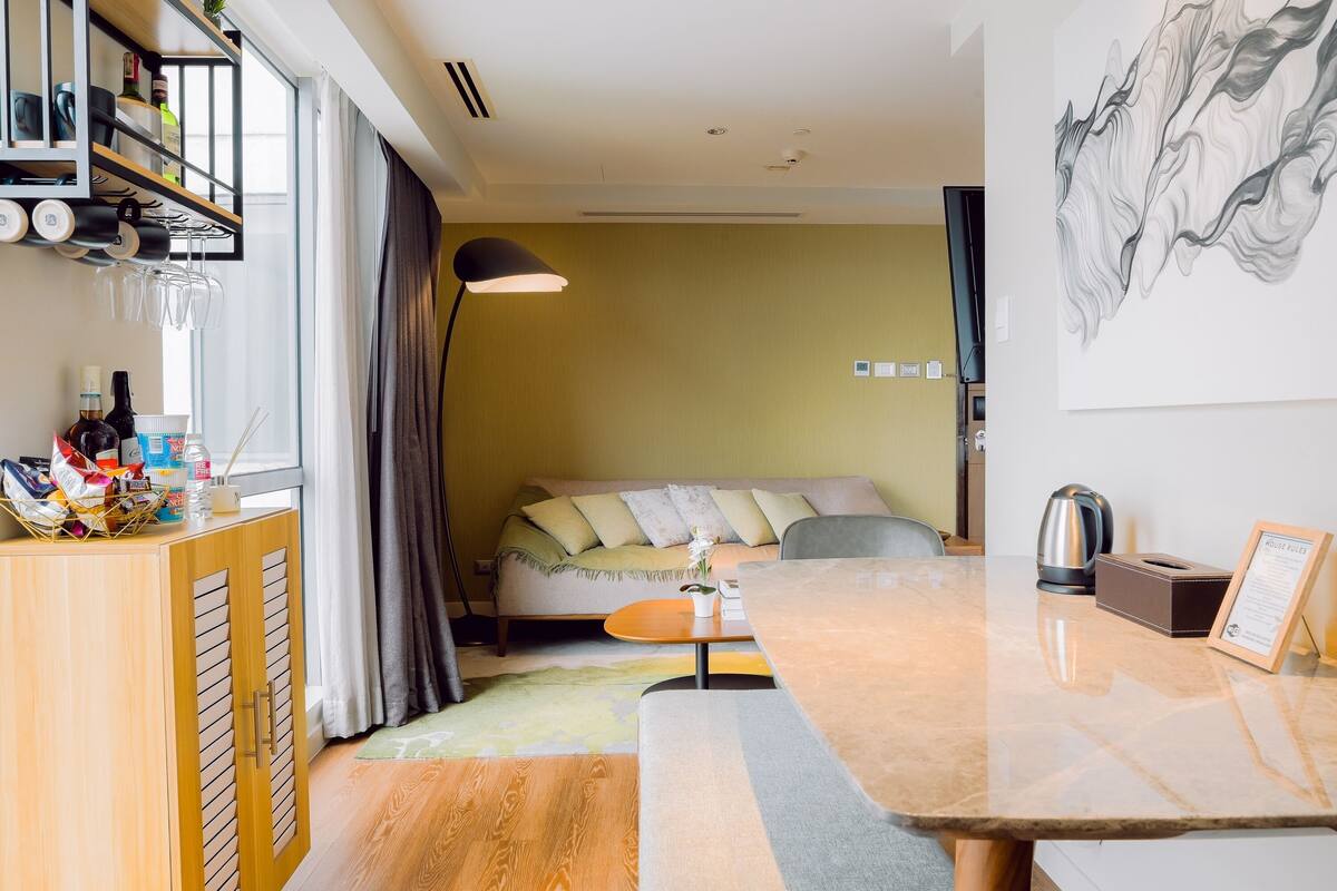 Quarto de hotel com cama, escrivaninha, cadeira, luminária, quadro e janela com cortinas.