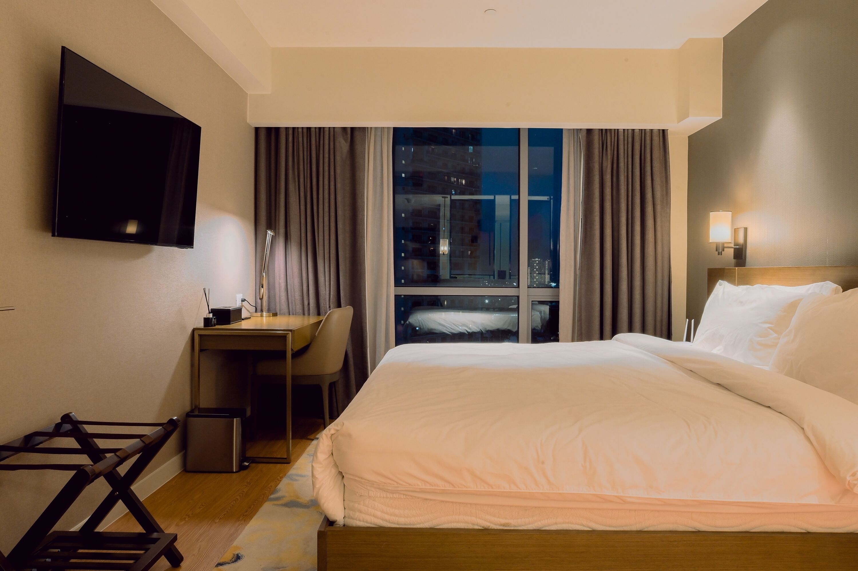 Quarto de hotel com cama, secretária, televisão e janela com cortinas.