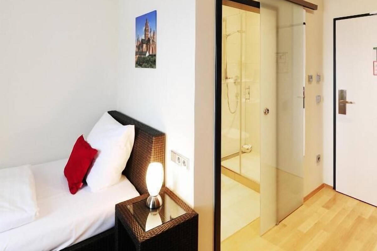 Quarto de hotel com uma cama, uma mesa de cabeceira com abajur, uma porta de vidro que dá para o banheiro e um quadro na parede.