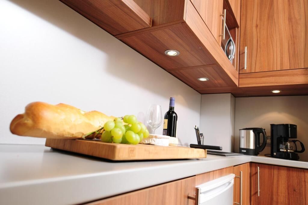 Cozinha com armários de madeira, tábua de corte com pão e uvas, garrafa de vinho e cafeteira.