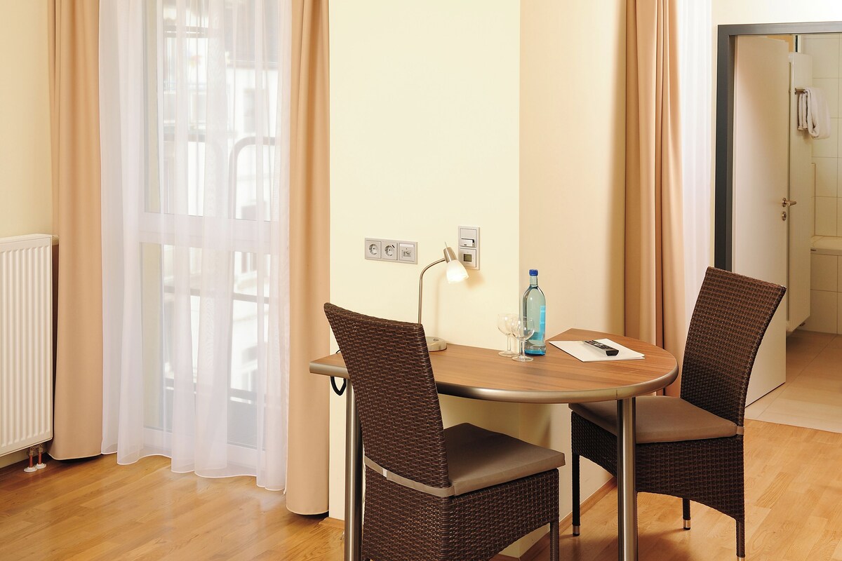 Quarto de hotel com cama, mesa de jantar com cadeiras, abajur, garrafa e controle remoto.