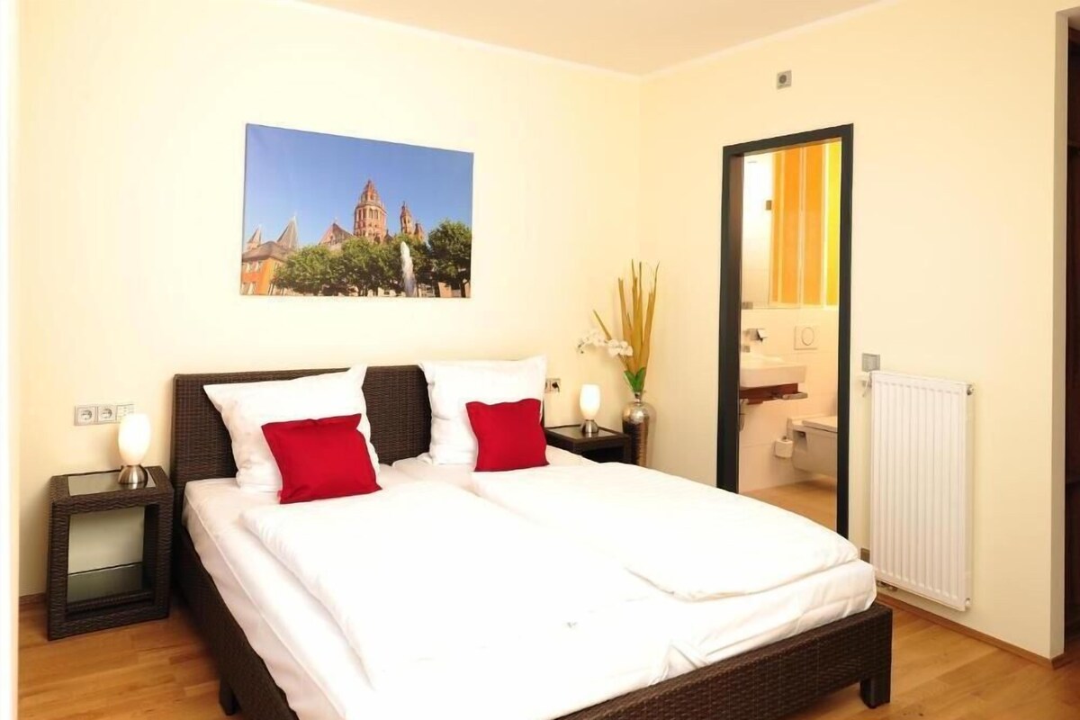 Cama bem arrumada com roupa de cama branca e vermelha, mesa de cabeceira com abajur, quadro na parede e vista de um banheiro por uma porta aberta.