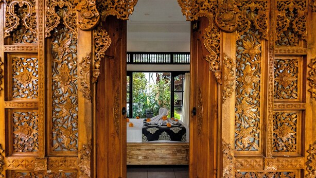 Uma porta de madeira ornamentada, com esculturas detalhadas, que dá acesso a um quarto com cama e vista para o verde lá fora.