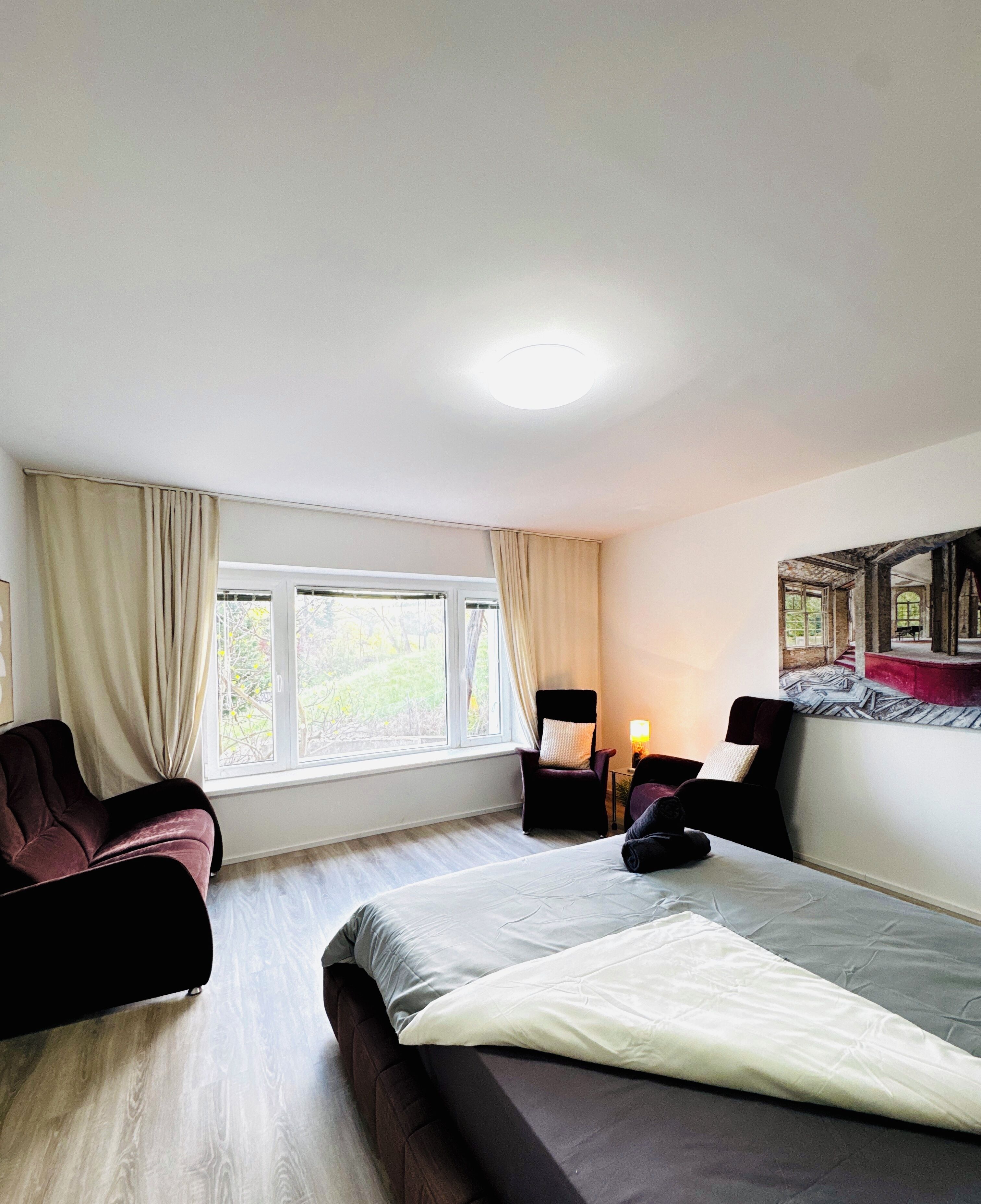 Quarto com cama, duas poltronas, janela com cortinas e uma obra de arte na parede.