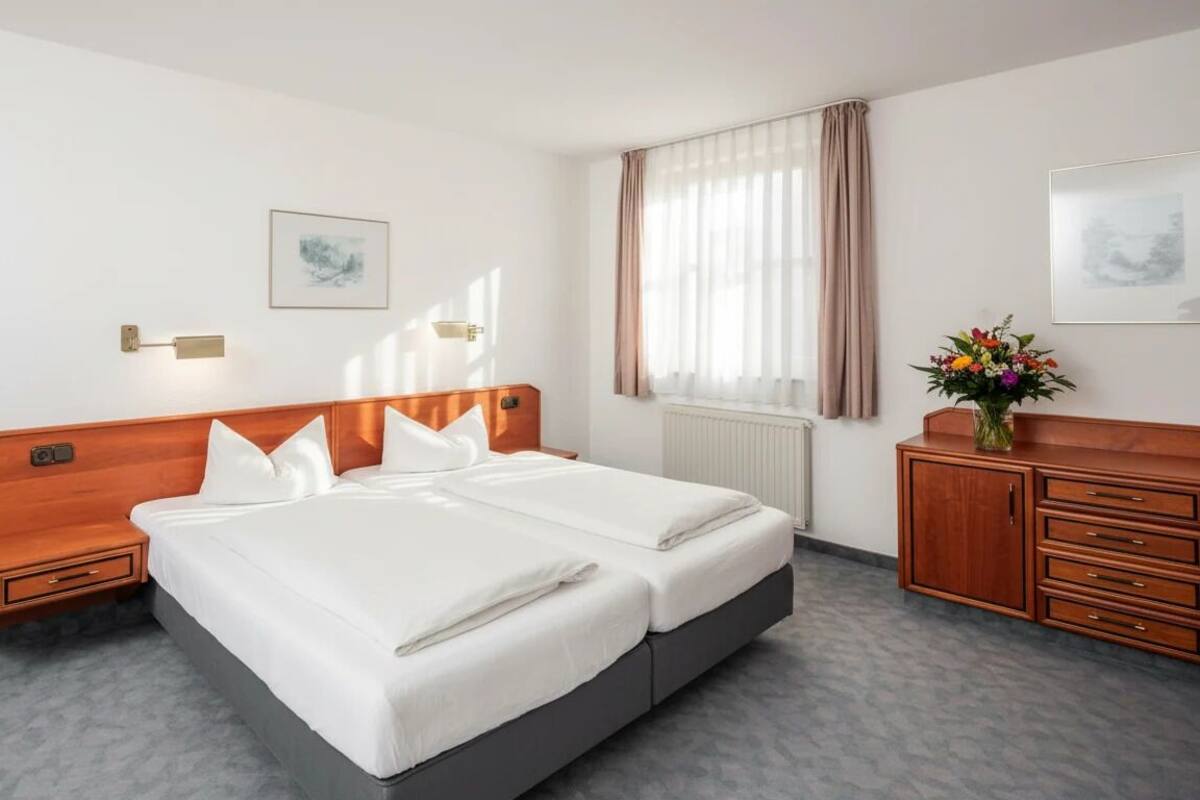 Uma cama de casal com cabeceira de madeira, mesa de cabeceira, cômoda e uma janela com cortinas.