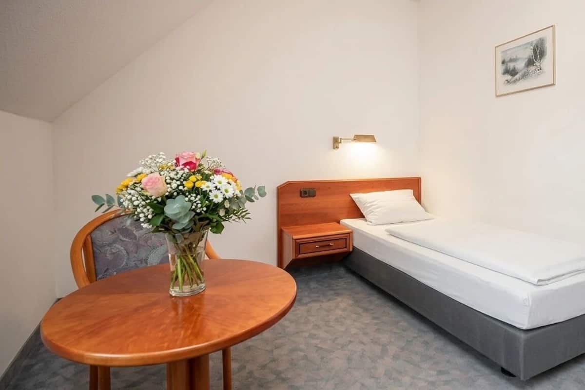 Uma cama de solteiro com cabeceira de madeira, uma mesa de cabeceira com abajur, um criado-mudo de madeira, uma cadeira e um vaso com flores.