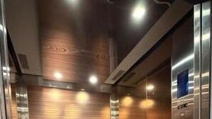 Interior de elevador com paredes metálicas, piso carpeteado e painel de comando à direita.