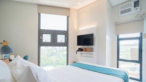 Quarto de hotel com cama, secretária com luminária, televisão e uma janela grande.