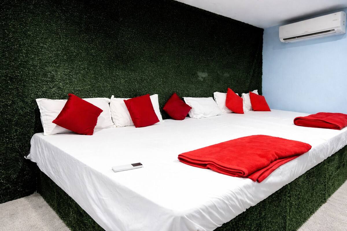 Quarto de hotel com uma cama grande, roupa de cama branca, almofadas vermelhas, uma mesa de cabeceira branca e um ar condicionado na parede.