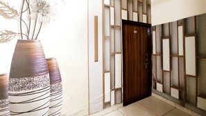 Um corredor com uma porta de madeira com a inscrição '102', um painel decorativo na parede e dois vasos com motivos florais.