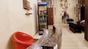 Uma área de recepção com balcão de mármore, uma cadeira vermelha, refrigerador de Pepsi e um painel decorativo com vasos.