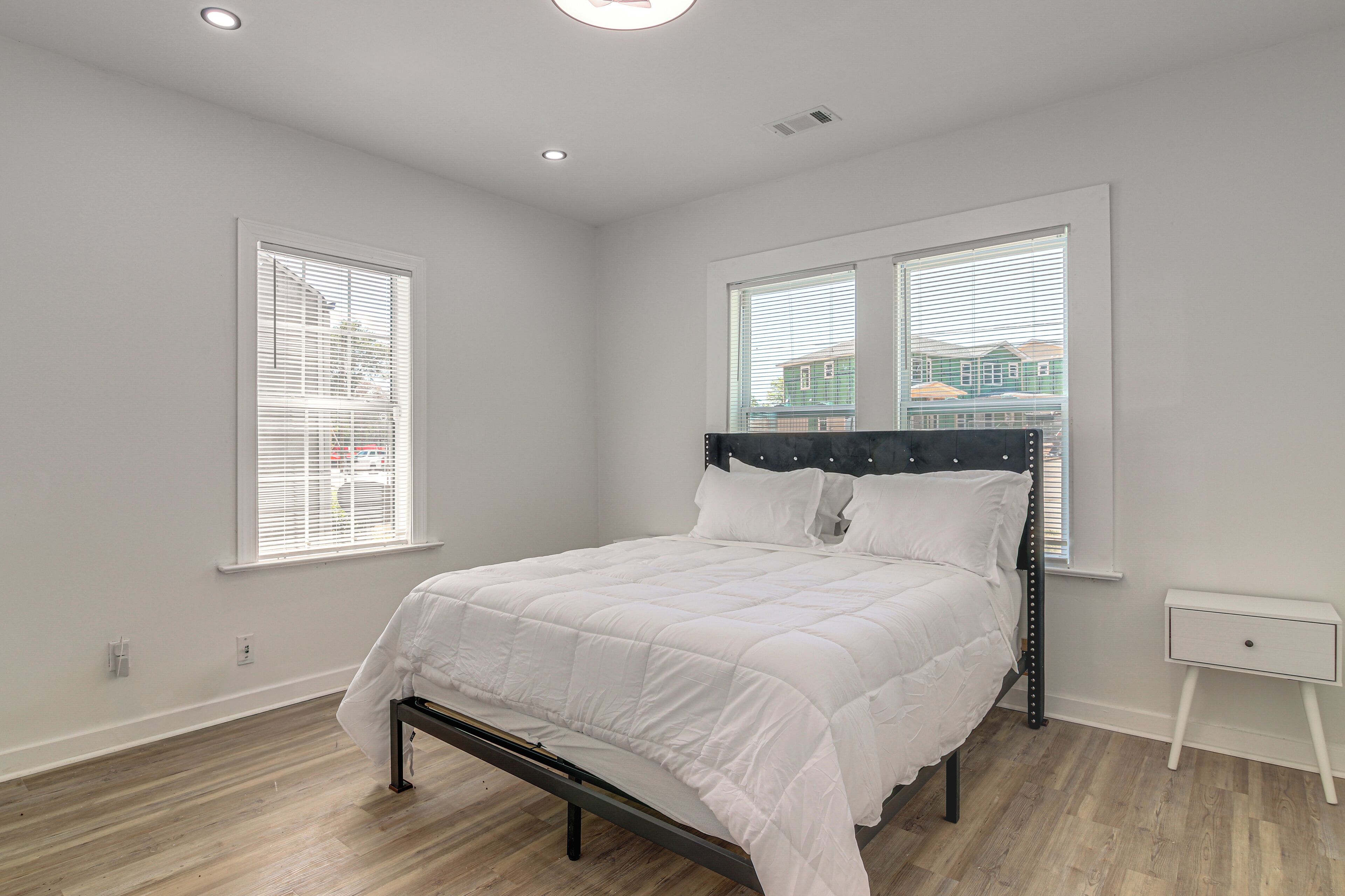 Quarto com uma cama, duas janelas, uma mesa de cabeceira e uma parede branca.