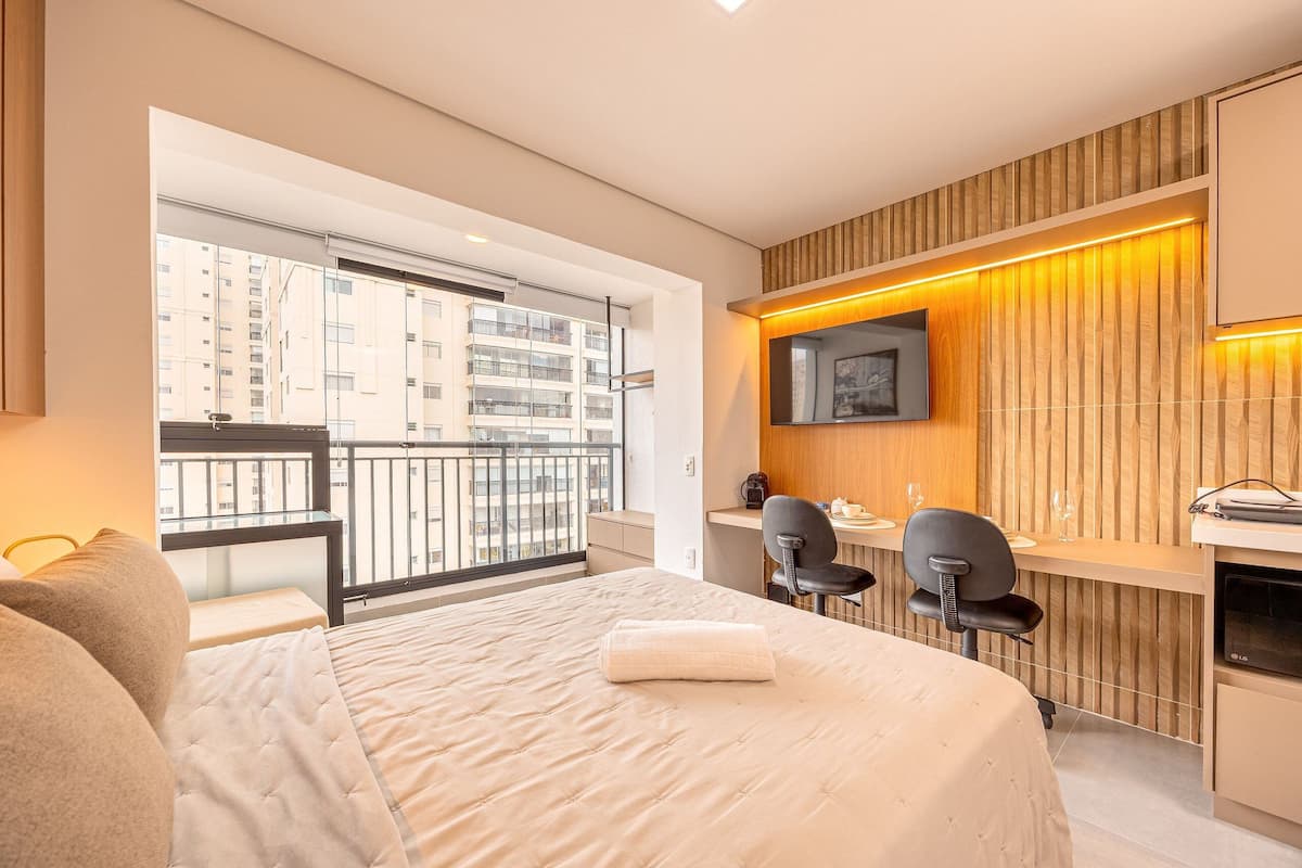 Quarto de hotel moderno com uma cama grande, uma escrivaninha com duas cadeiras, televisão e varanda com vista para a cidade.