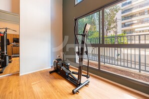 Uma academia com equipamentos de exercício, uma janela grande com vista para um edifício e árvores, e piso de madeira.