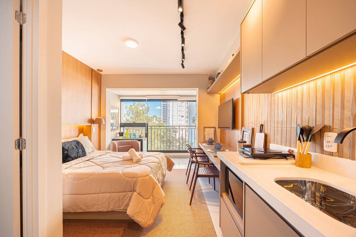 Quarto de hotel moderno com cama, cozinha compacta e varanda vista para a cidade.