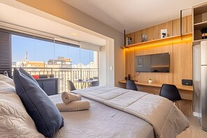 Quarto de hotel moderno com uma cama grande, varanda com vista para a cidade, televisão de tela plana na parede e uma mesa pequena com cadeiras.