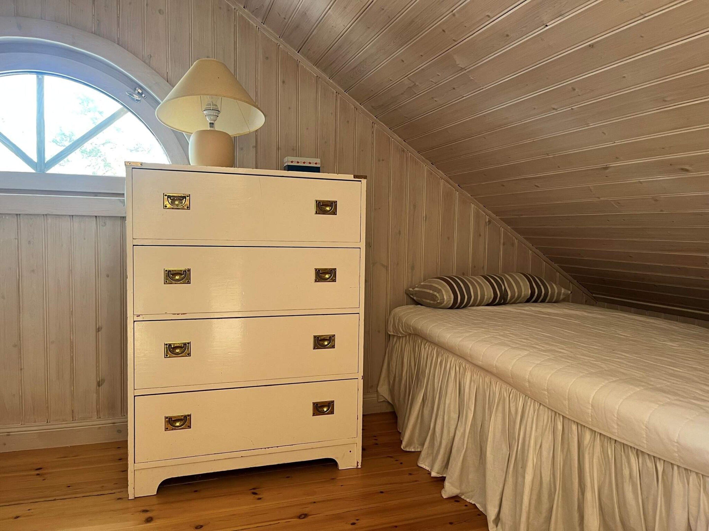 Quarto com teto de madeira, penteadeira com gavetas, cama com travesseiro listrado e janela com vista.