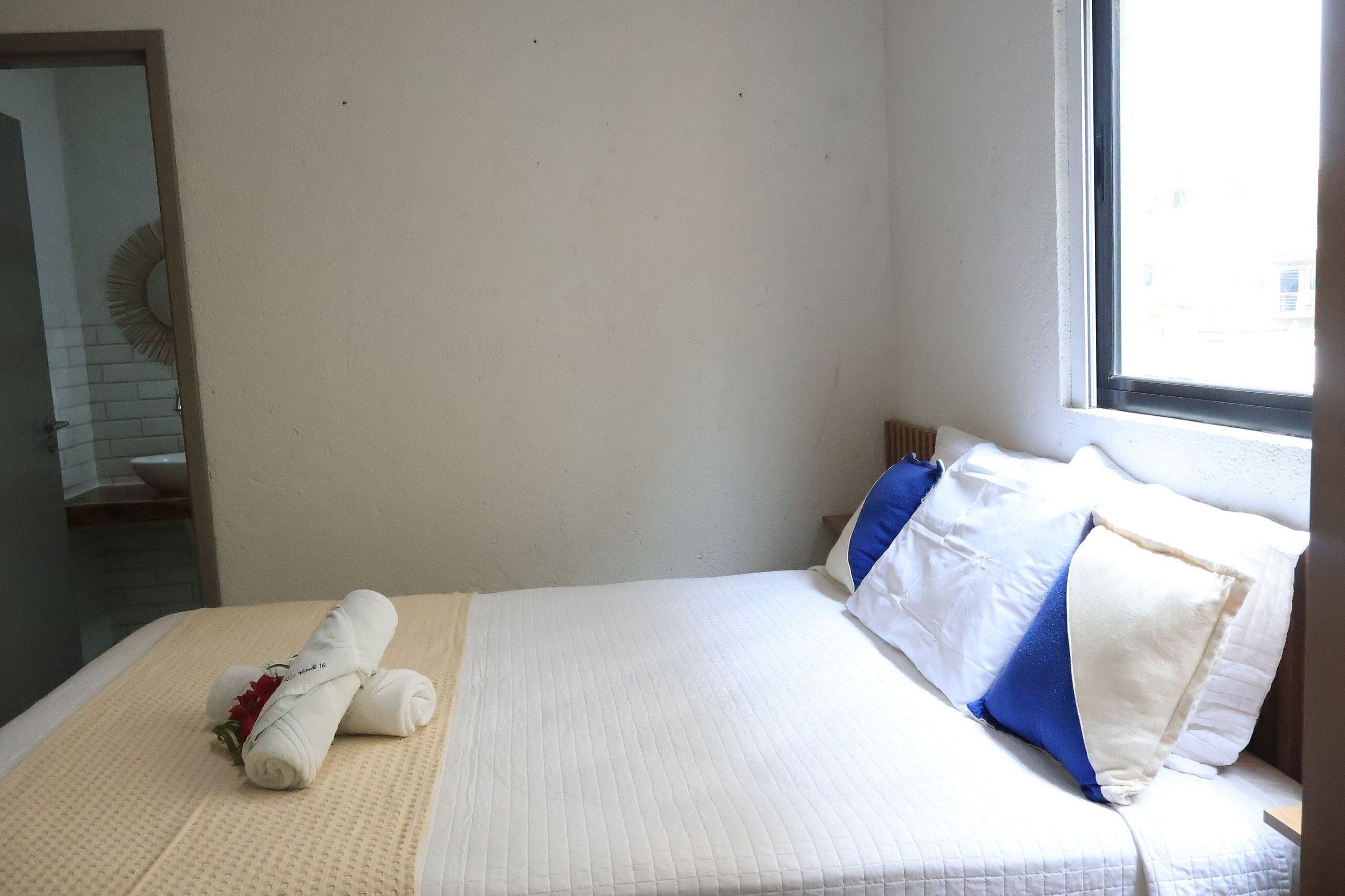 Uma cama bem arrumada com colcha branca e almofadas azuis, uma janela com vista e uma porta que dá para outro quarto.