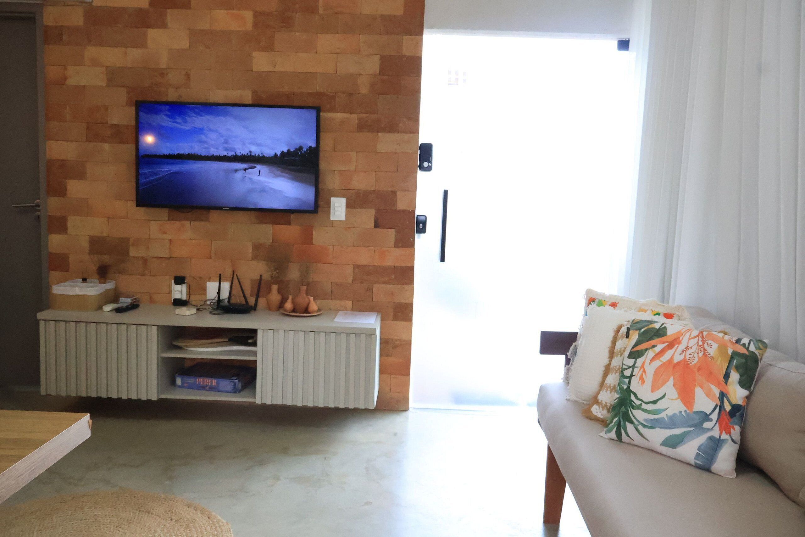 Uma sala de estar com uma televisão fixada numa parede de tijolos, um sofá com almofada florida e um radiador branco abaixo.