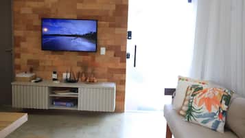 Uma sala de estar com uma televisão fixada numa parede de tijolos, um sofá com almofada florida e um radiador branco abaixo.